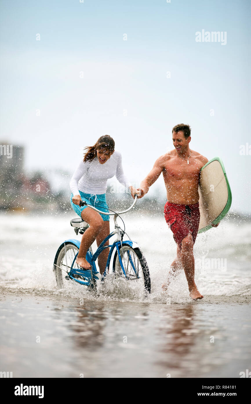 Man with surfboard running alongside a woman on a bike at the beach ...
