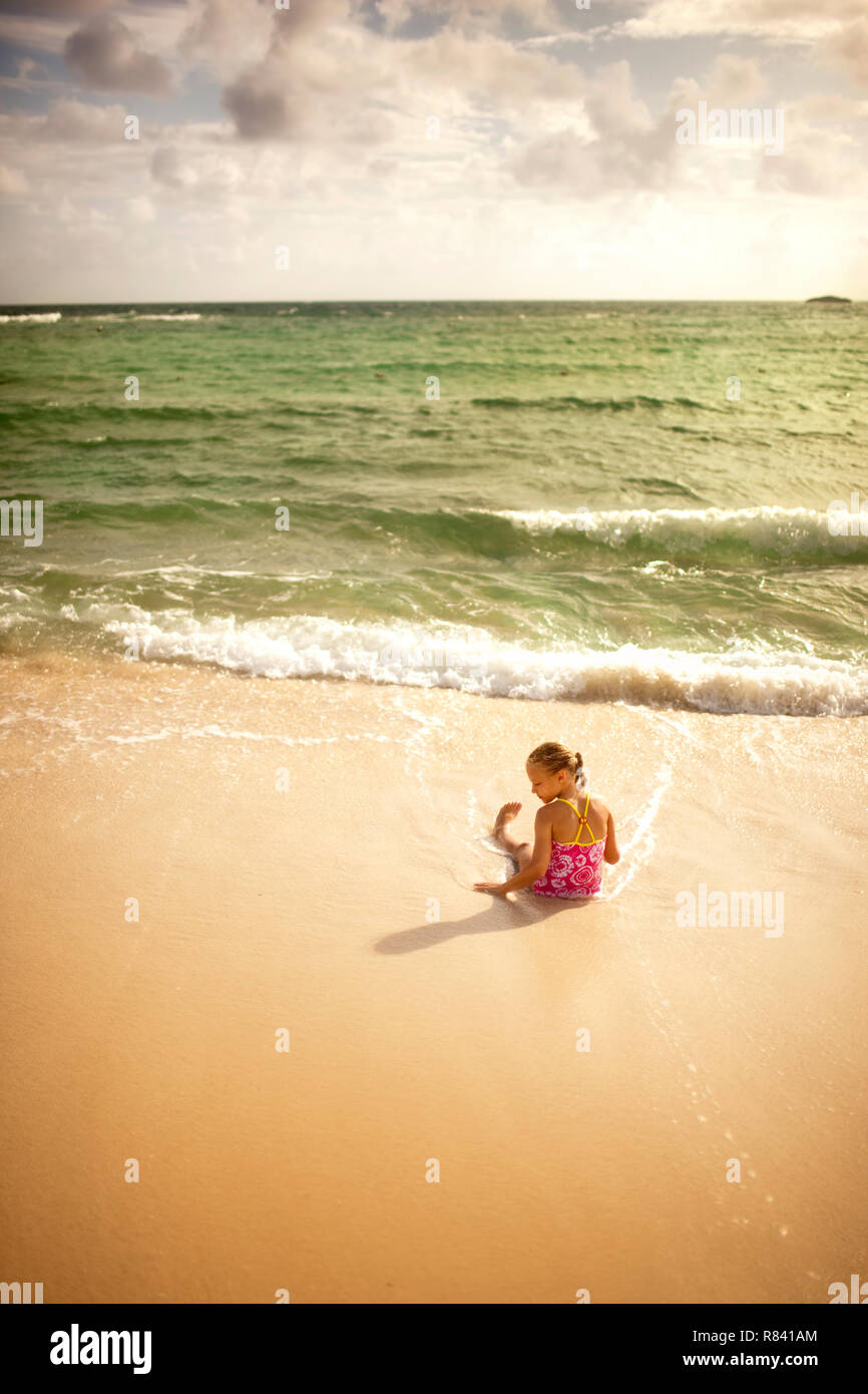 Young Girl Sitting Alone At Waters Edge On A Sandy Beach While Wearing