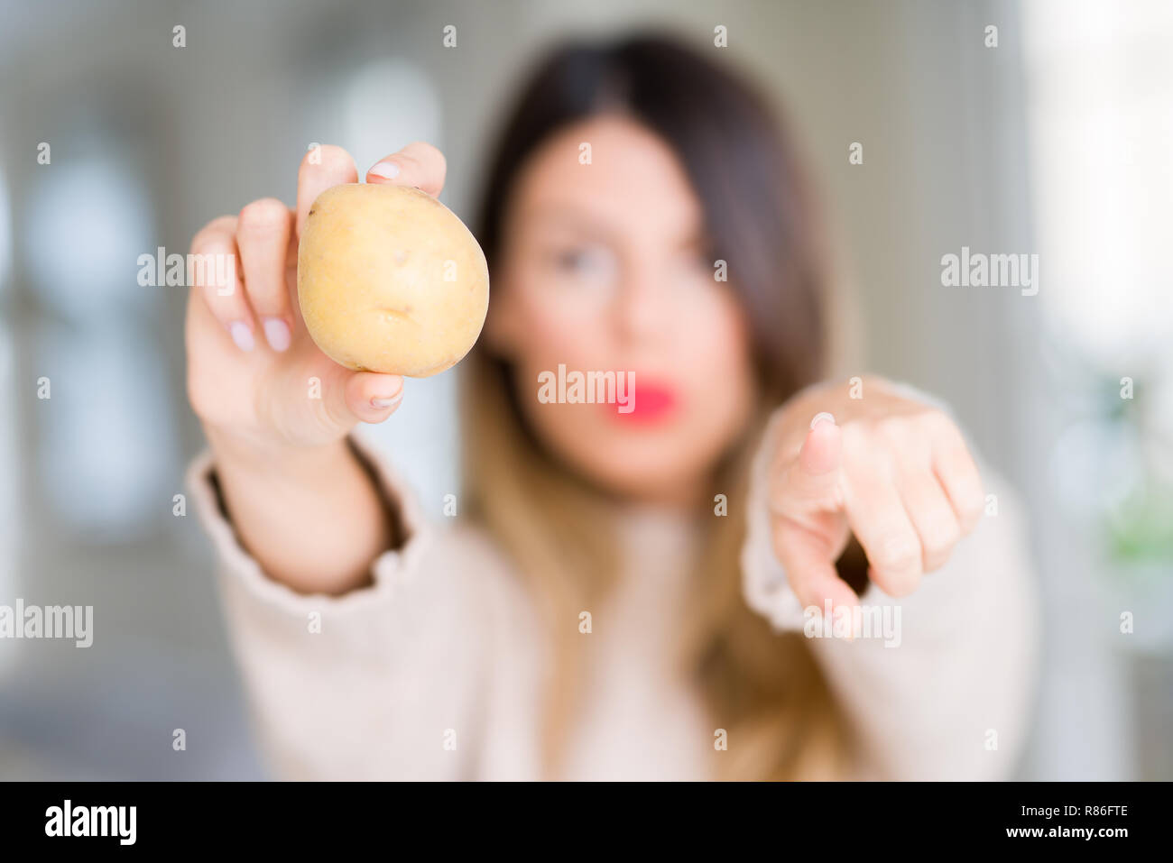 Young beautiful woman holding fresh potato at home pointing with finger ...