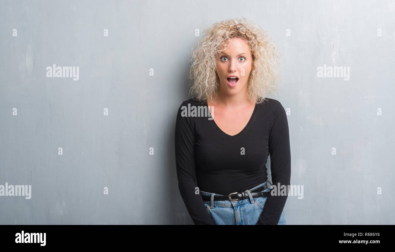 Young blonde woman with curly hair over grunge grey background scared ...