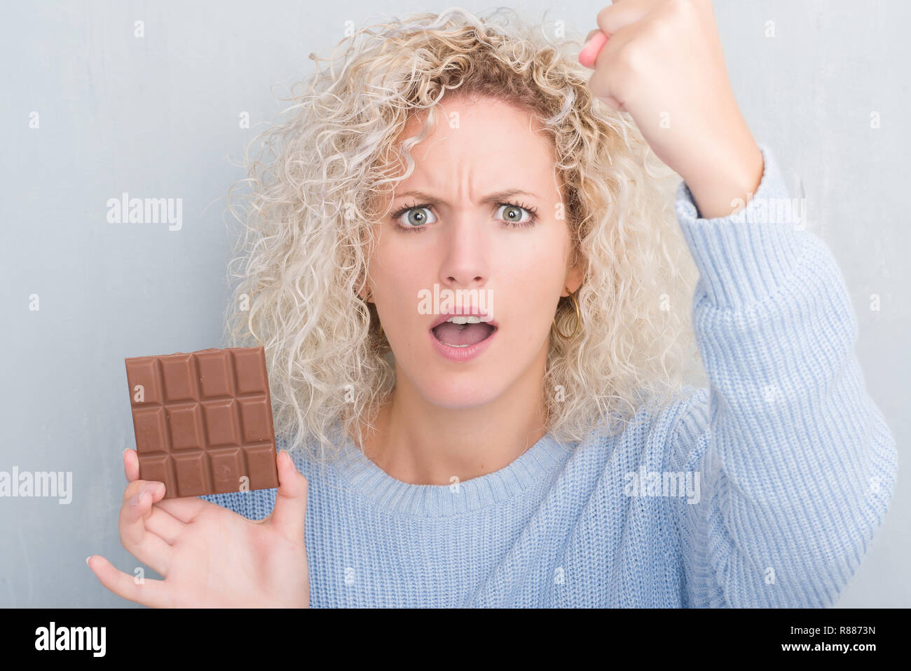 Young blonde woman over grunge grey wall eating chocolate bar annoyed ...