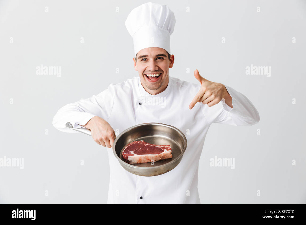 Cheerful chef cook wearing uniform showing frying pan with a raw beef ...
