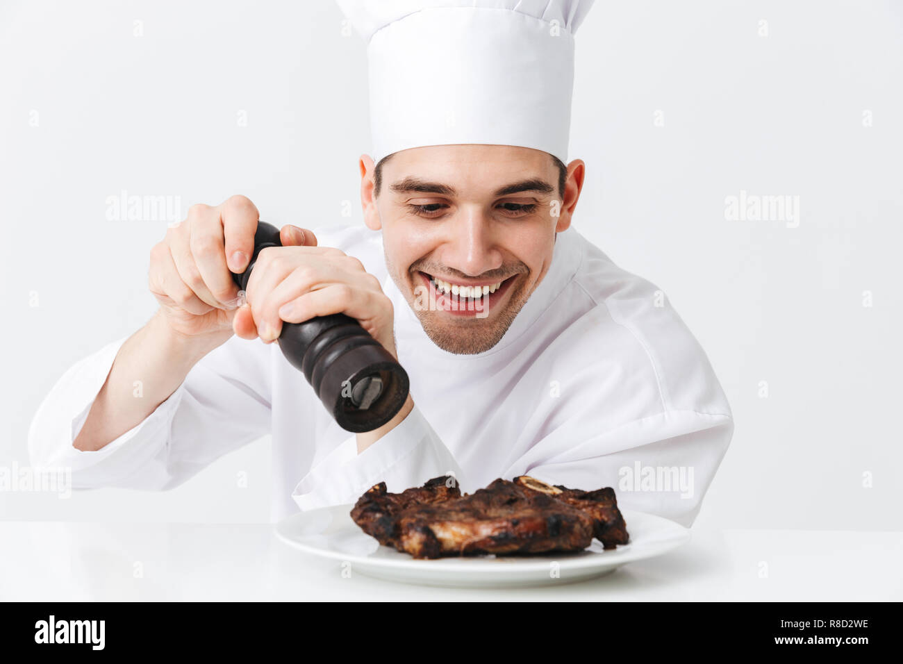 Cheerful chef cook wearing uniform peppers cooked beef steak on a plate ...