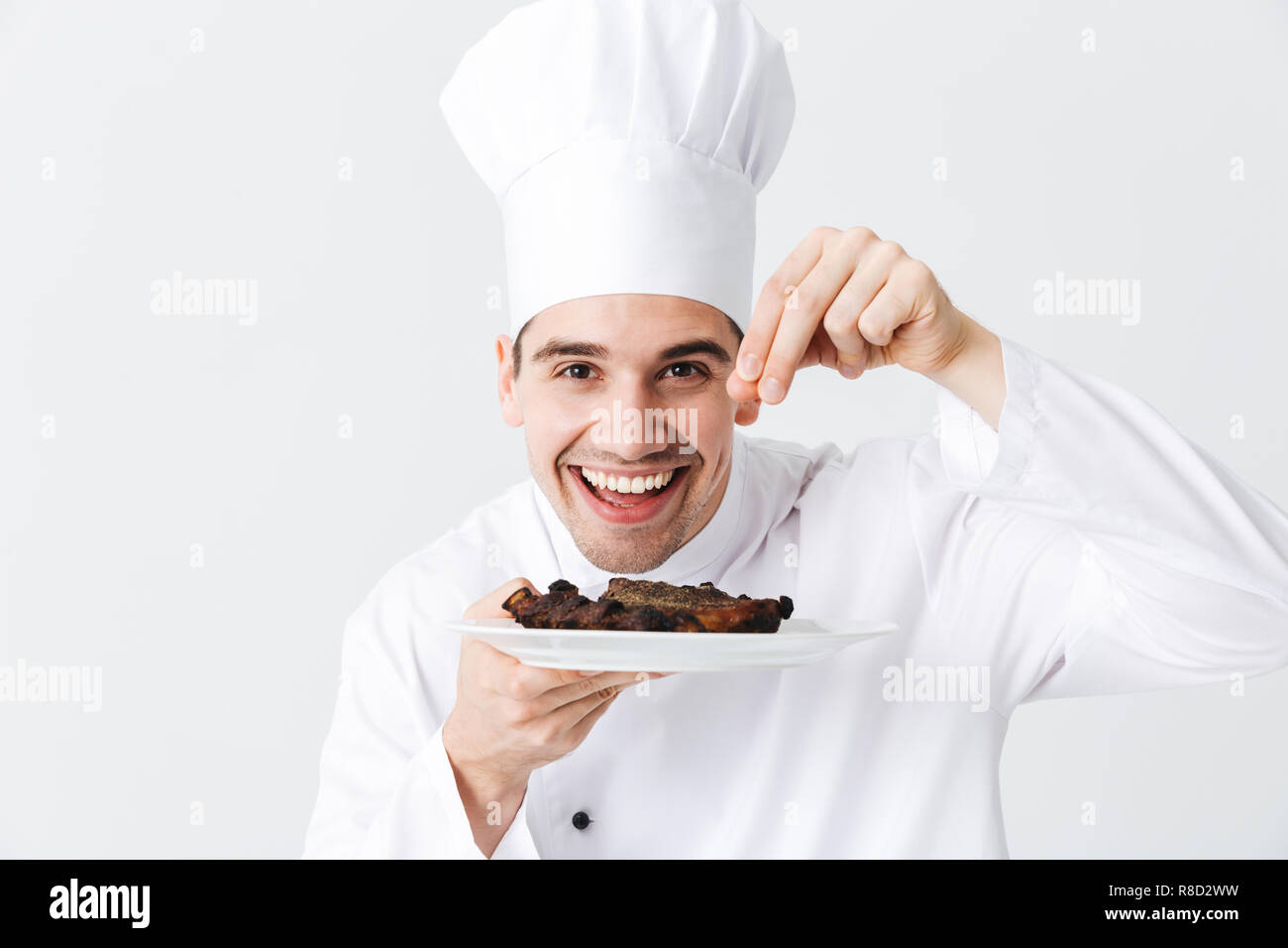 Cheerful chef cook wearing uniform peppers cooked beef steak on a plate ...