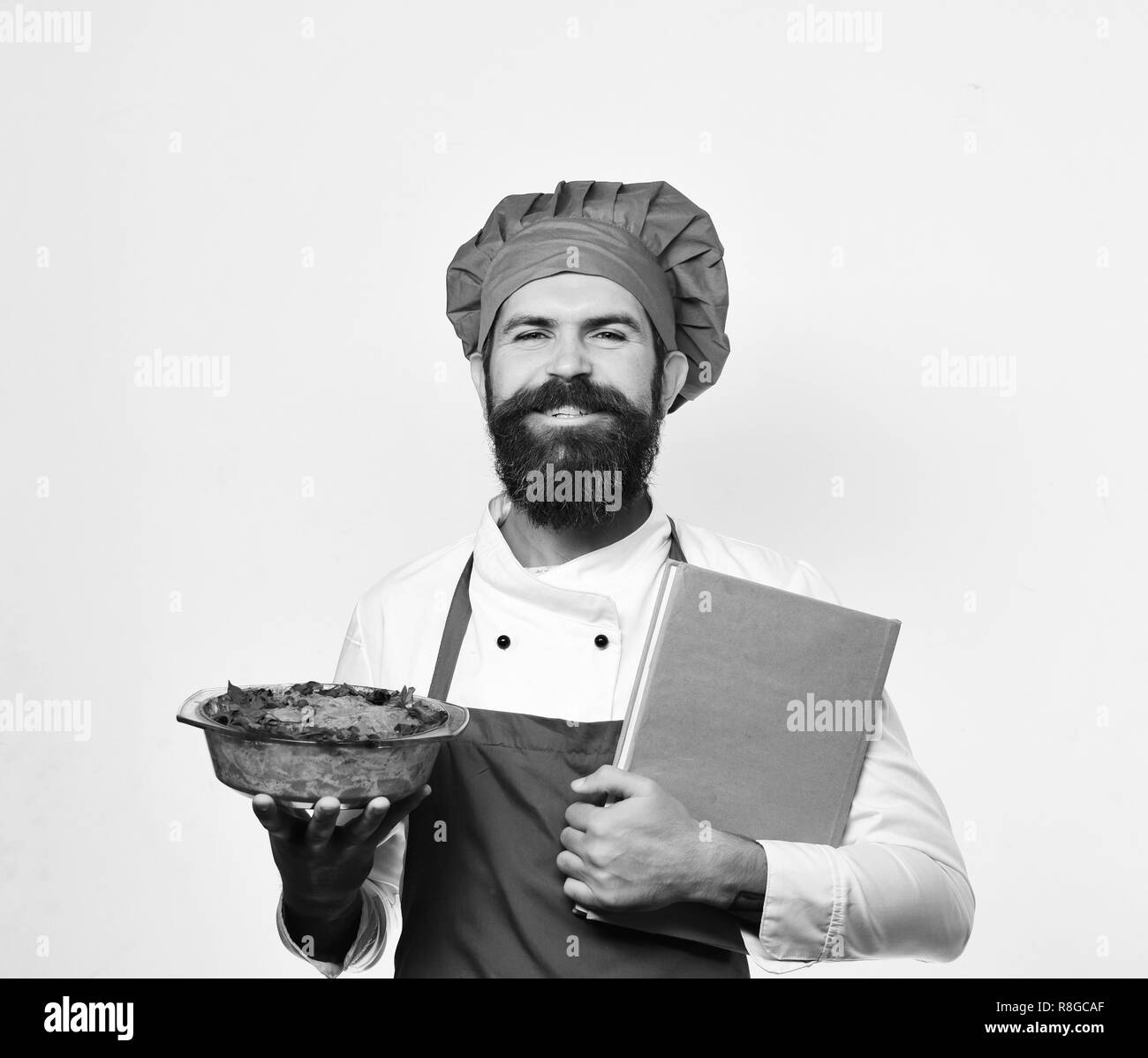Cook with proud smile in burgundy uniform holds baked dish and book ...