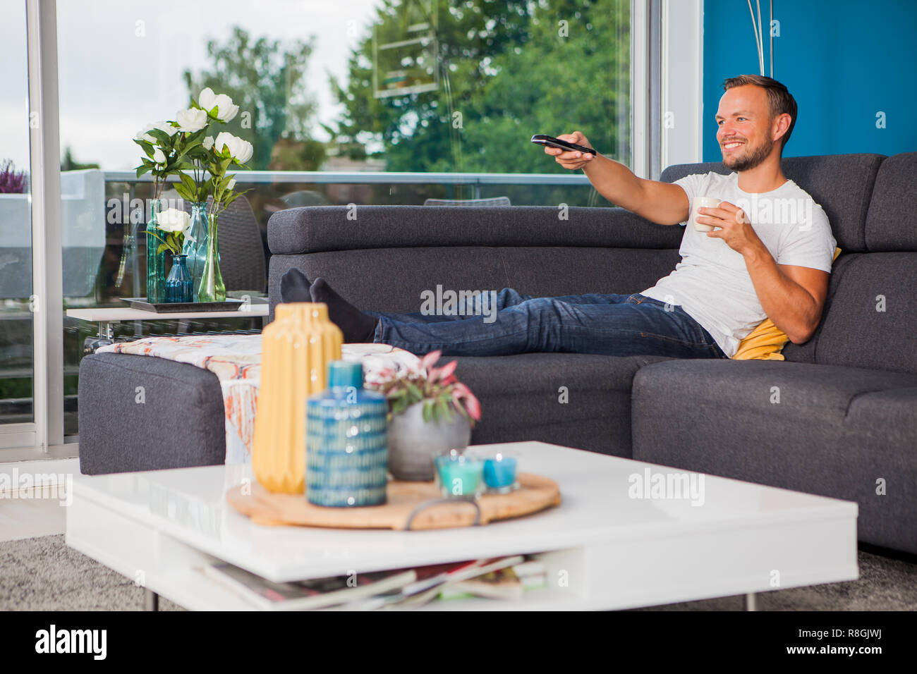 Young man using a remote control while drinking coffee on the couch in ...