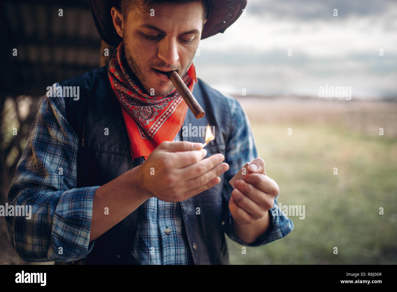 Bearded cowboy lights a cigar with matches, texas ranch on background ...