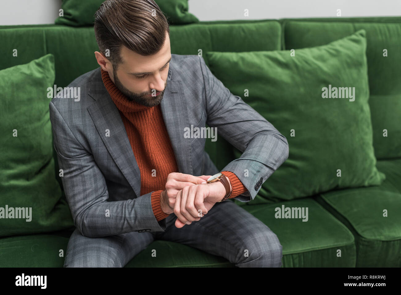 man in formal wear sitting on green sofa and adjusting watch on hand ...