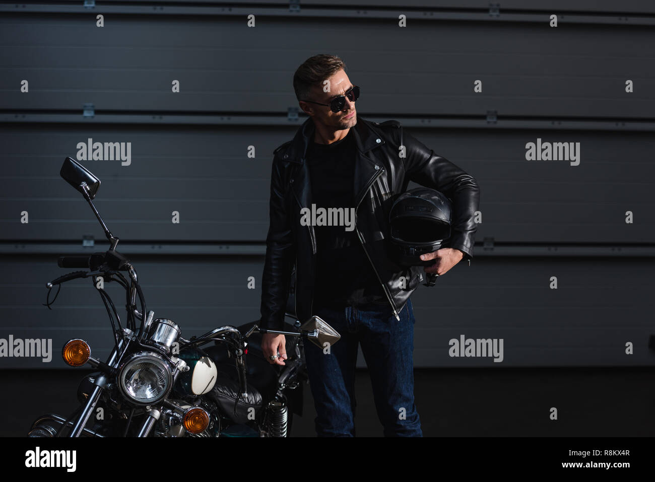 handsome biker in sunglasses holding helment and standing by motorcycle ...