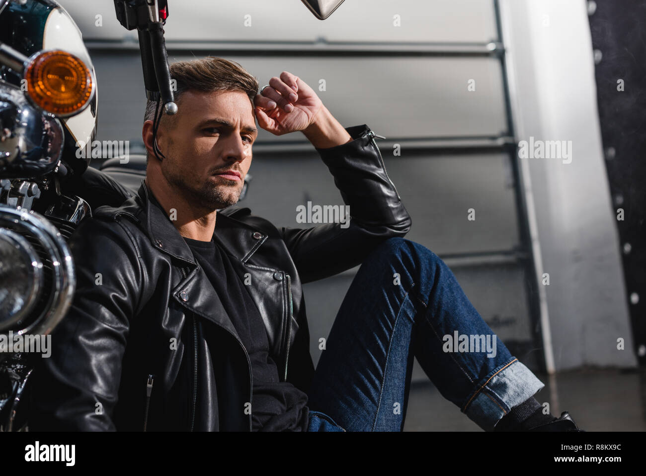 handsome biker looking at camera and leaning on motorbike in garage ...