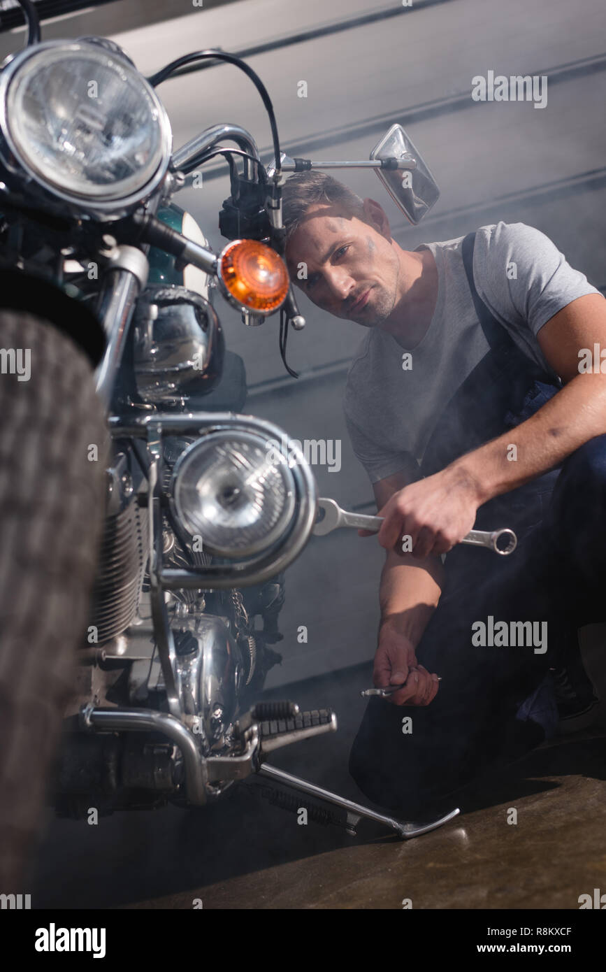 handsome mechanic holding wrench and fixing motorcycle engine in garage ...
