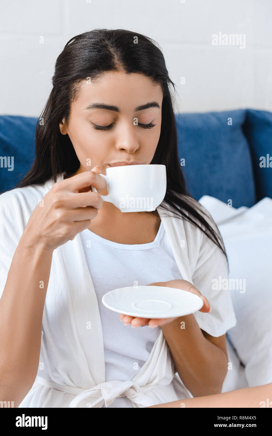 attractive mixed race girl in white robe drinking coffee in bed in ...