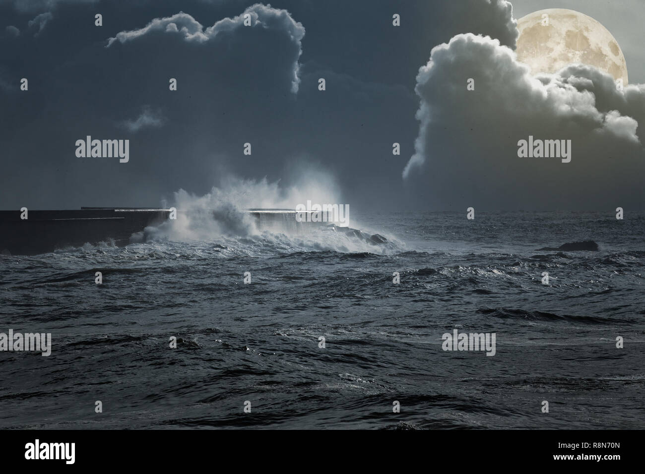 Big ocean wave breaking against harbor wall in a cloudy full moon night ...
