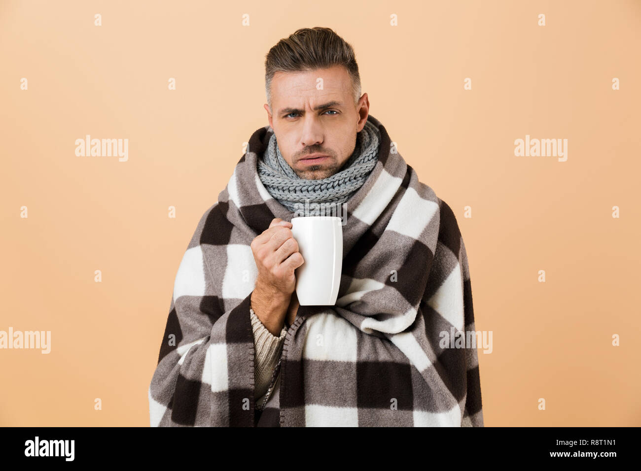 Portrait of a sad sick man wrapped in a blanket standing isolated over ...