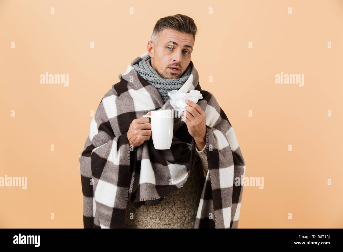 Portrait of a sad sick man wrapped in a blanket standing isolated over ...