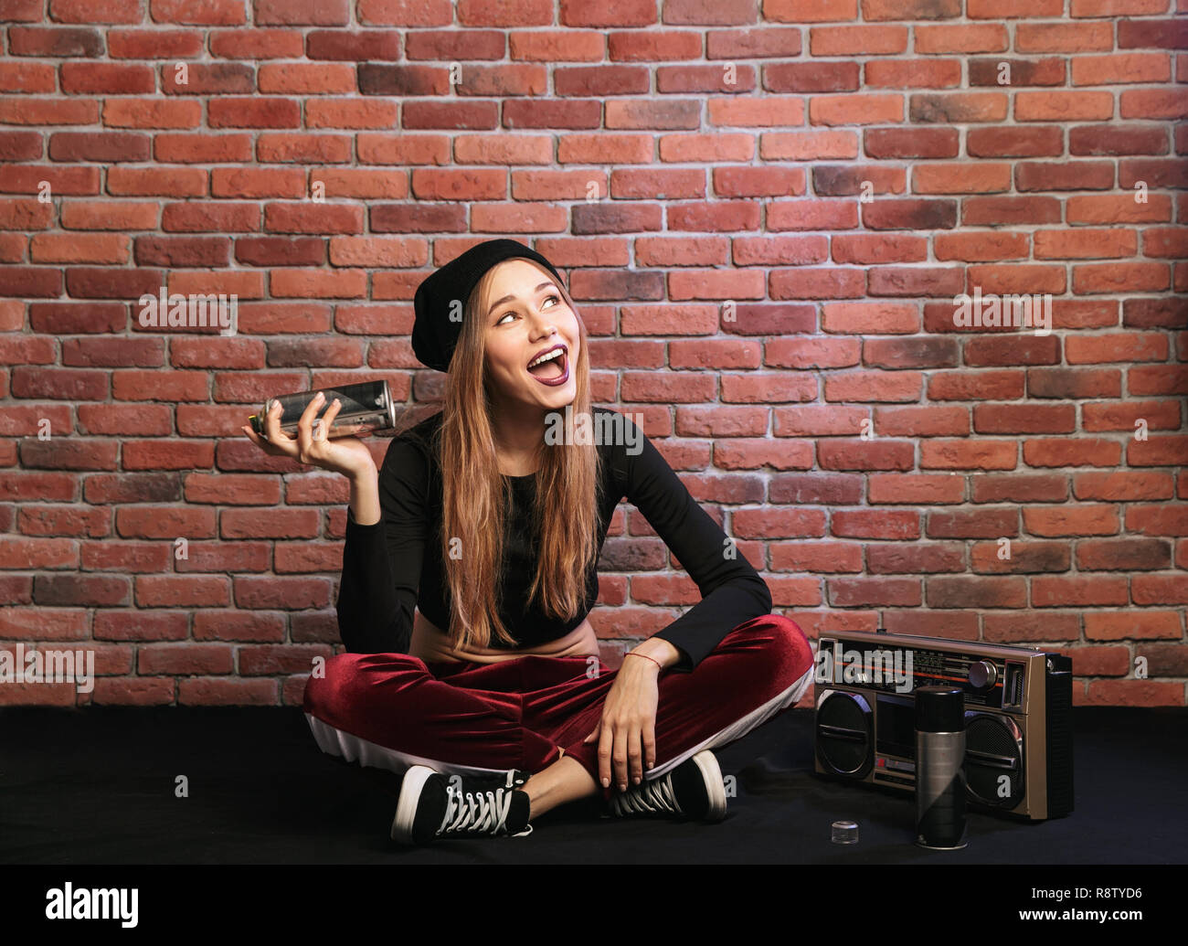 Photo of joyous young hip hop girl sitting on floor against brick wall ...