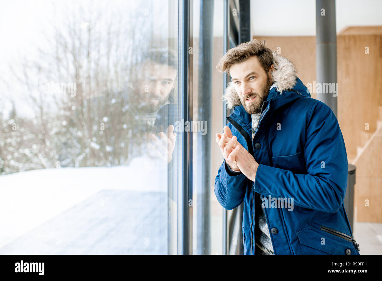 Man dressed in winter clothes feeling cold standing near the window at ...