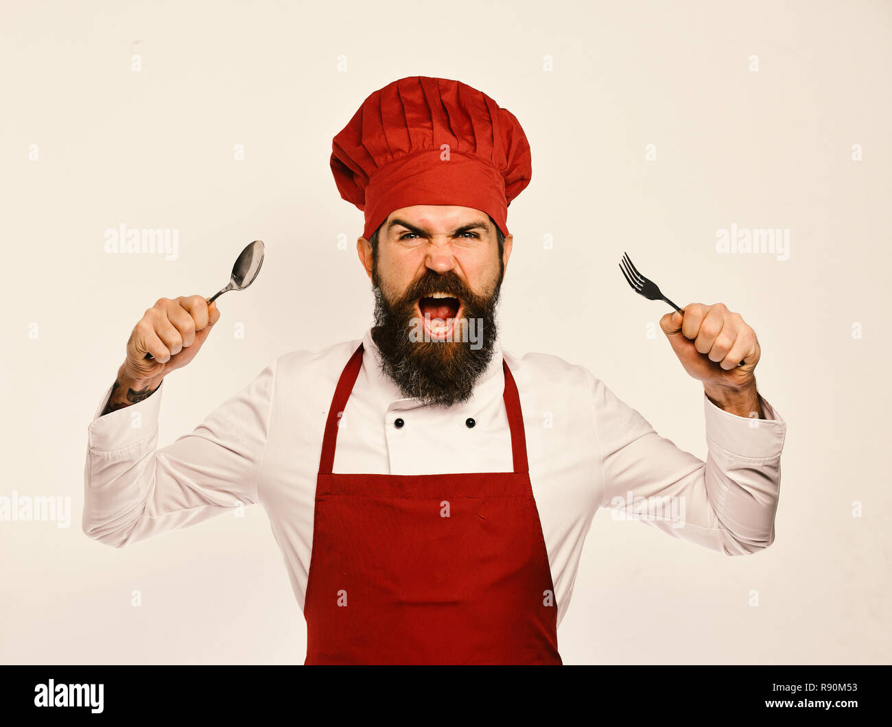Cook with angry face in burgundy uniform shows cutlery. Chef presents ...
