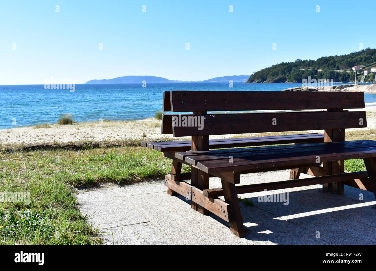 Bench on a beach promenade with grass and vegetation in sand. Blue sea ...