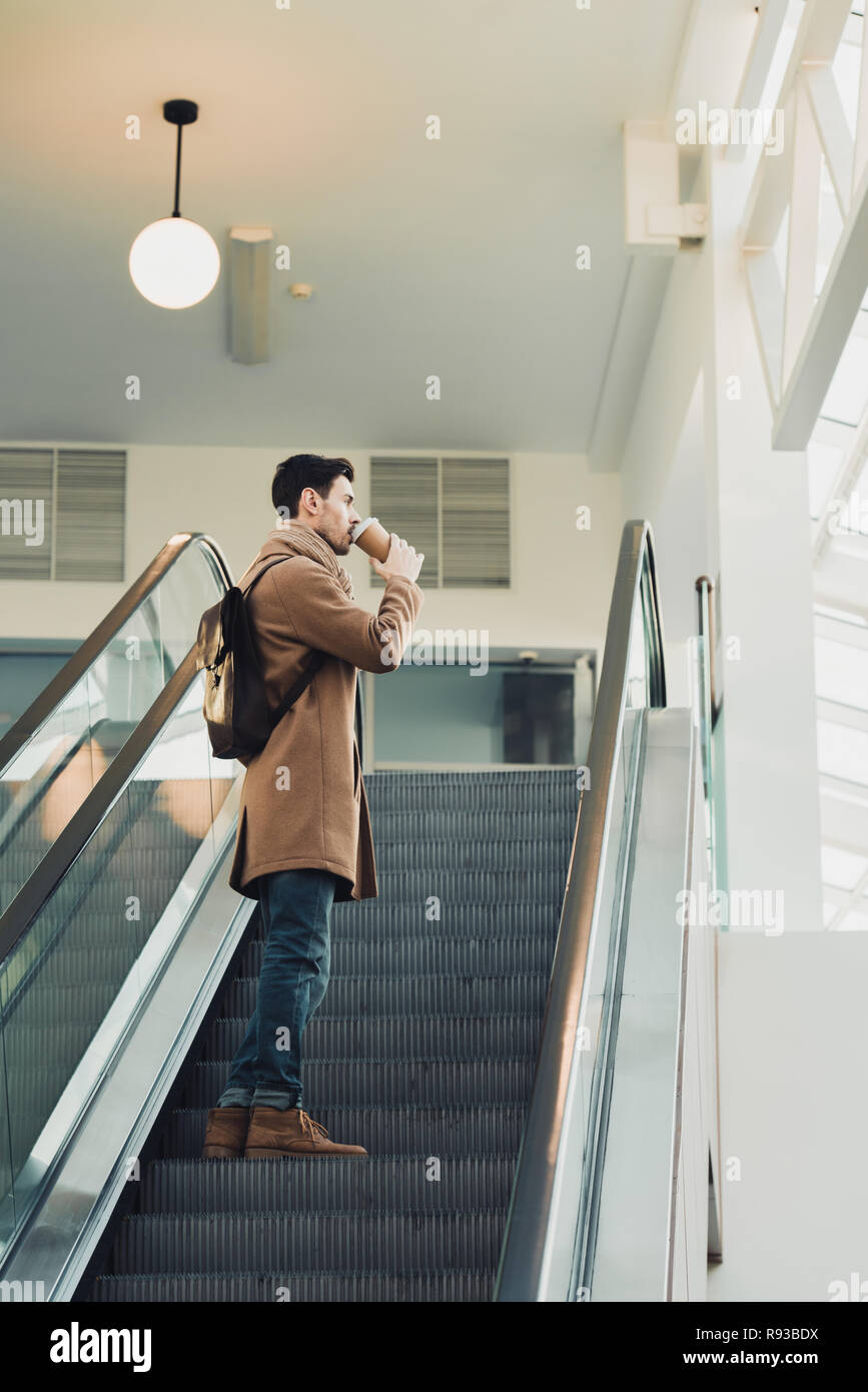 handsome man going up on escalator and drinking from disposable cup ...