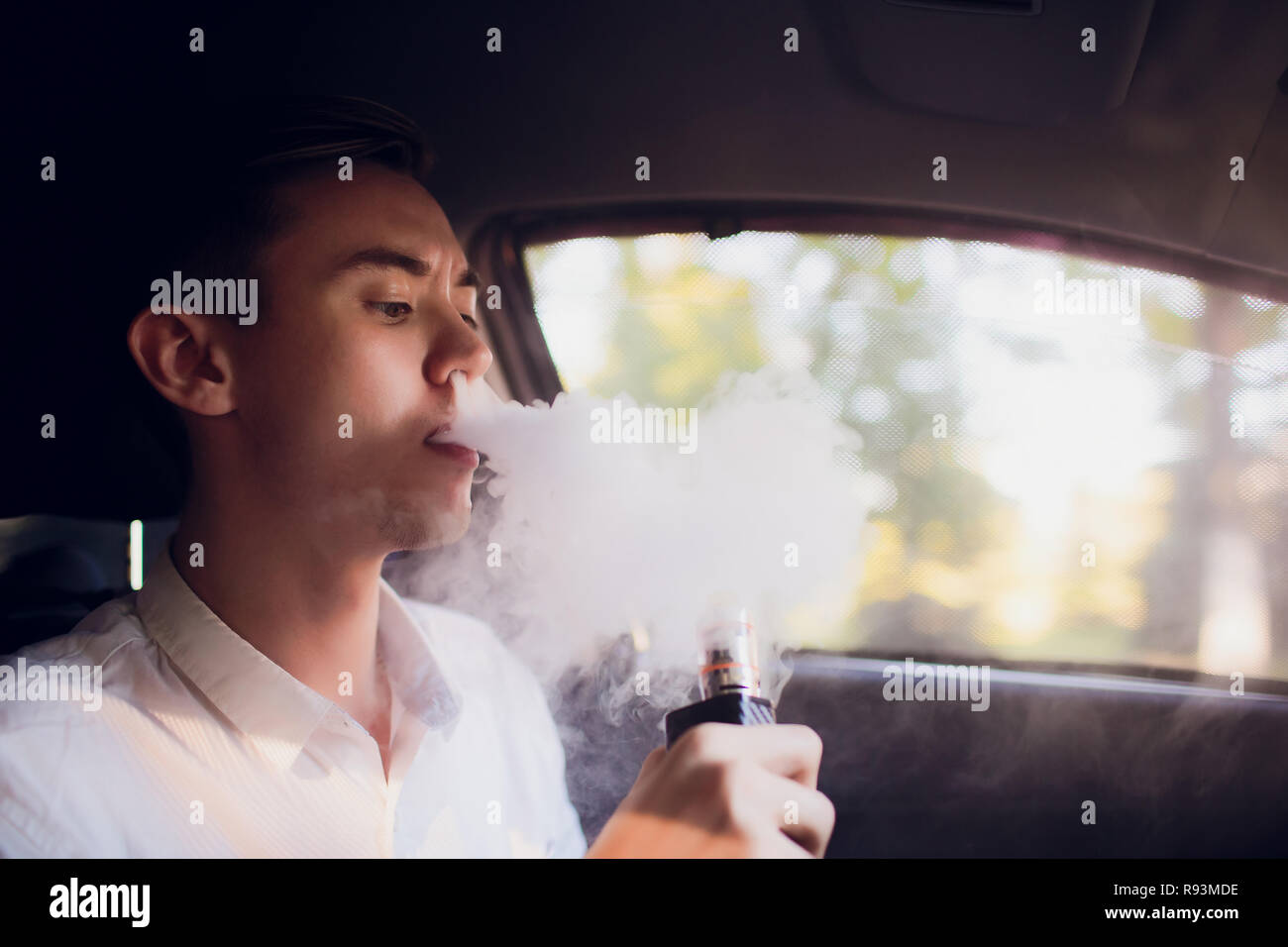 View from the side of a young man smoking an e-cigarette as he drives ...