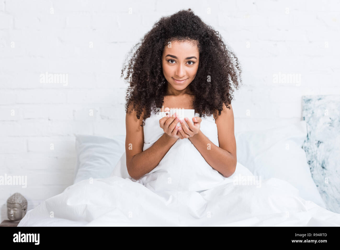 pretty curly african american girl holding cup of coffee in bed during ...