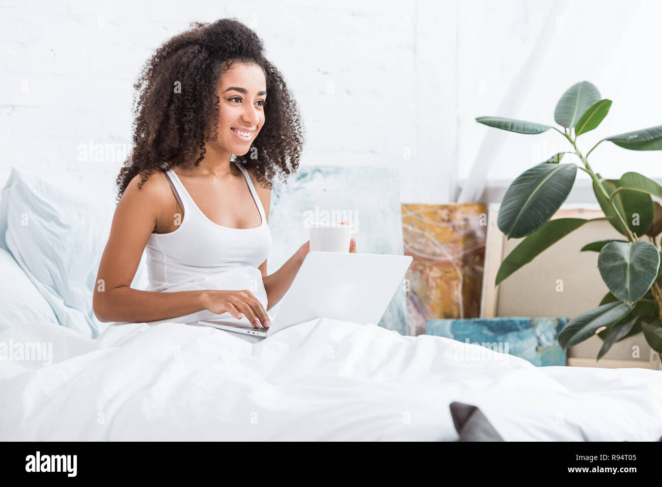 young woman holding coffee cup and using laptop in bed during morning ...