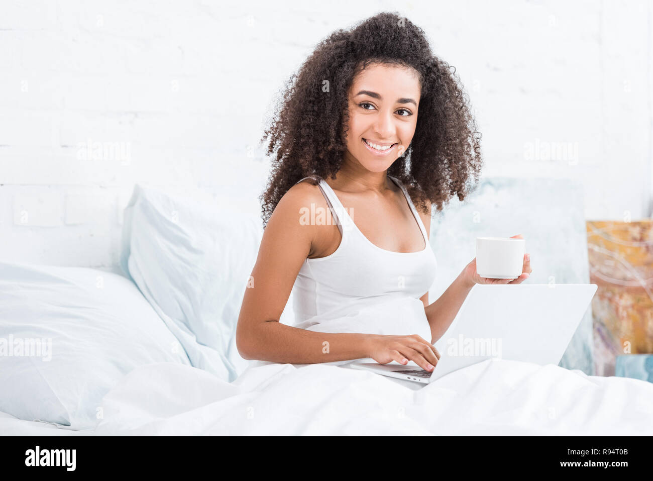 joyful curly girl holding coffee cup and using laptop in bed during ...