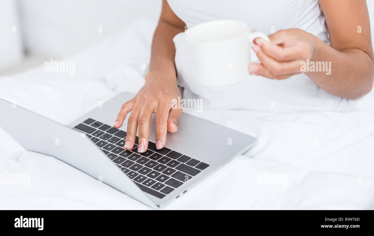 cropped image of woman with coffee cup using laptop in bed during ...