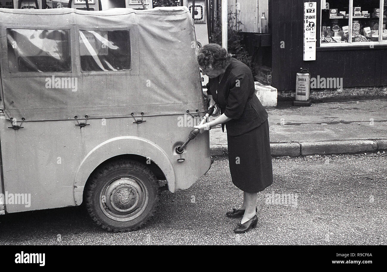 1955, historical, in a rural high street, a lady holding a petrol pump ...