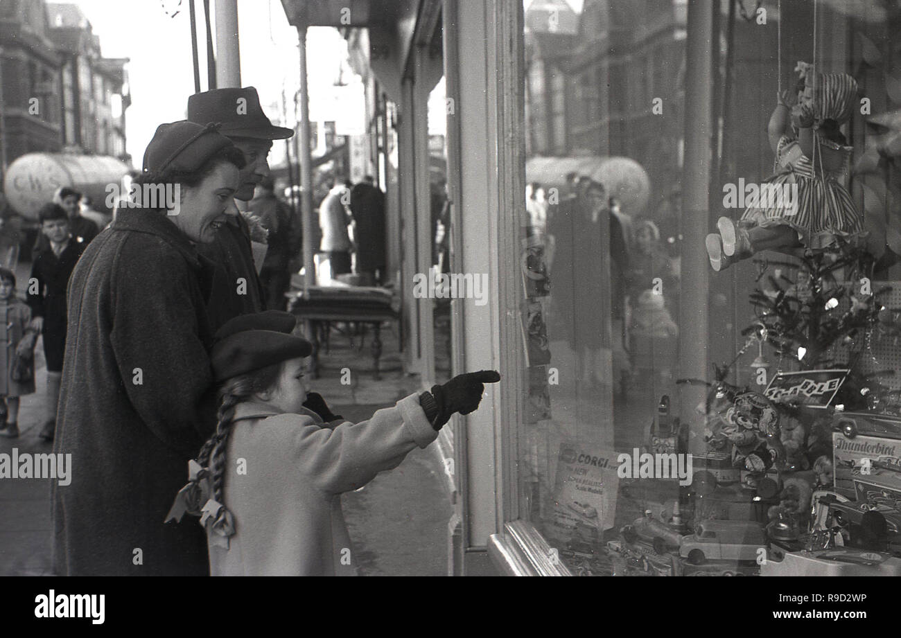 1950s, historical, a child with her parents in a street looking into a ...