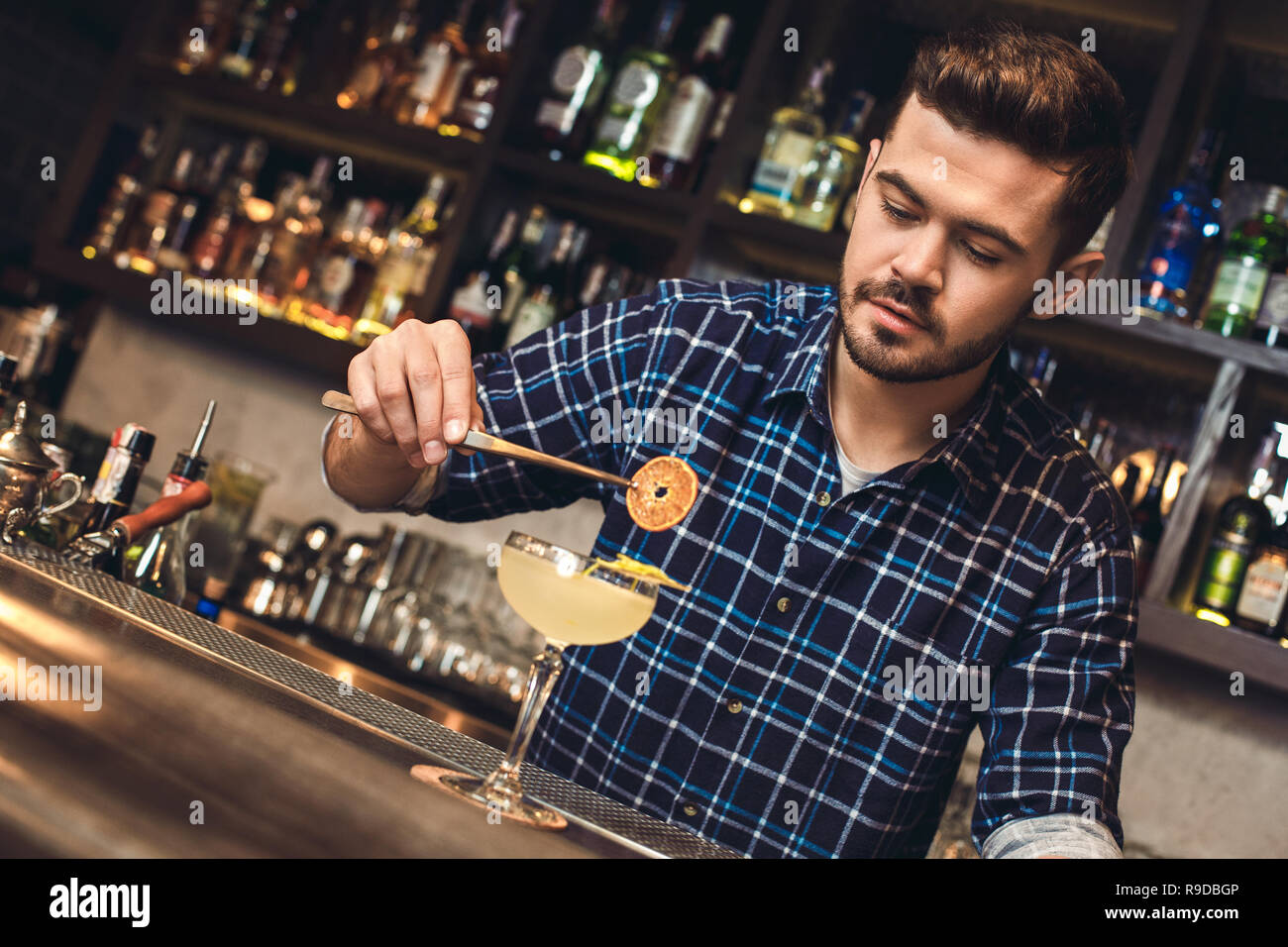 Young barman standing at bar counter decorating glass of cocktail with ...