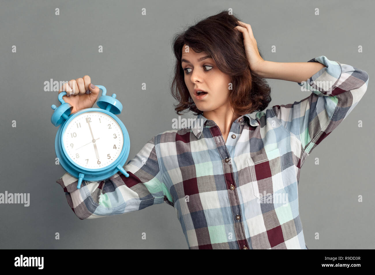Young woman standing isolated on grey wall holding alarm clock looking ...