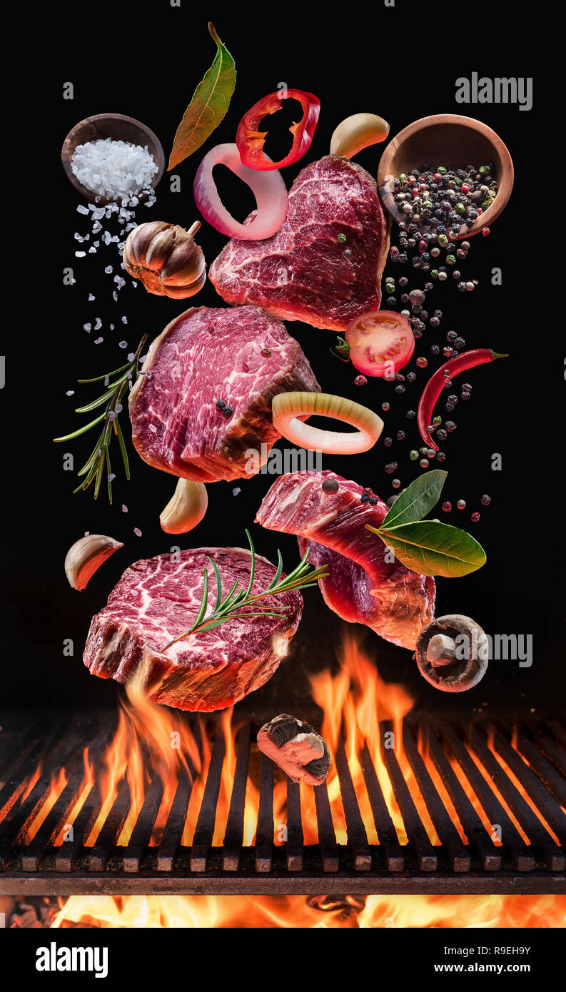 Raw beef steaks with vegetables and spices fly over the blazing grill ...
