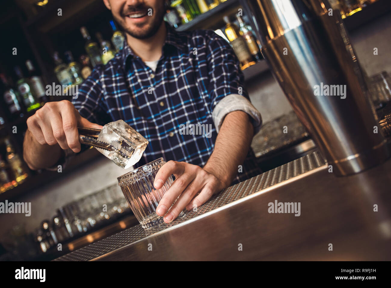 Young barman standing at bar counter putting ice cube into glass ...