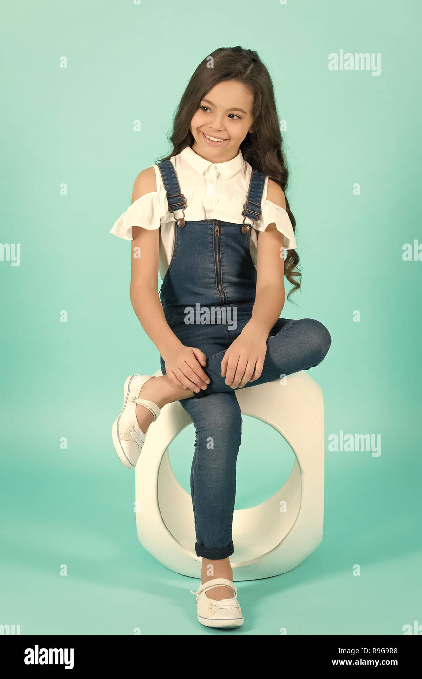 Child model in jeans overall sit leg crossed on chair. Girl with long ...