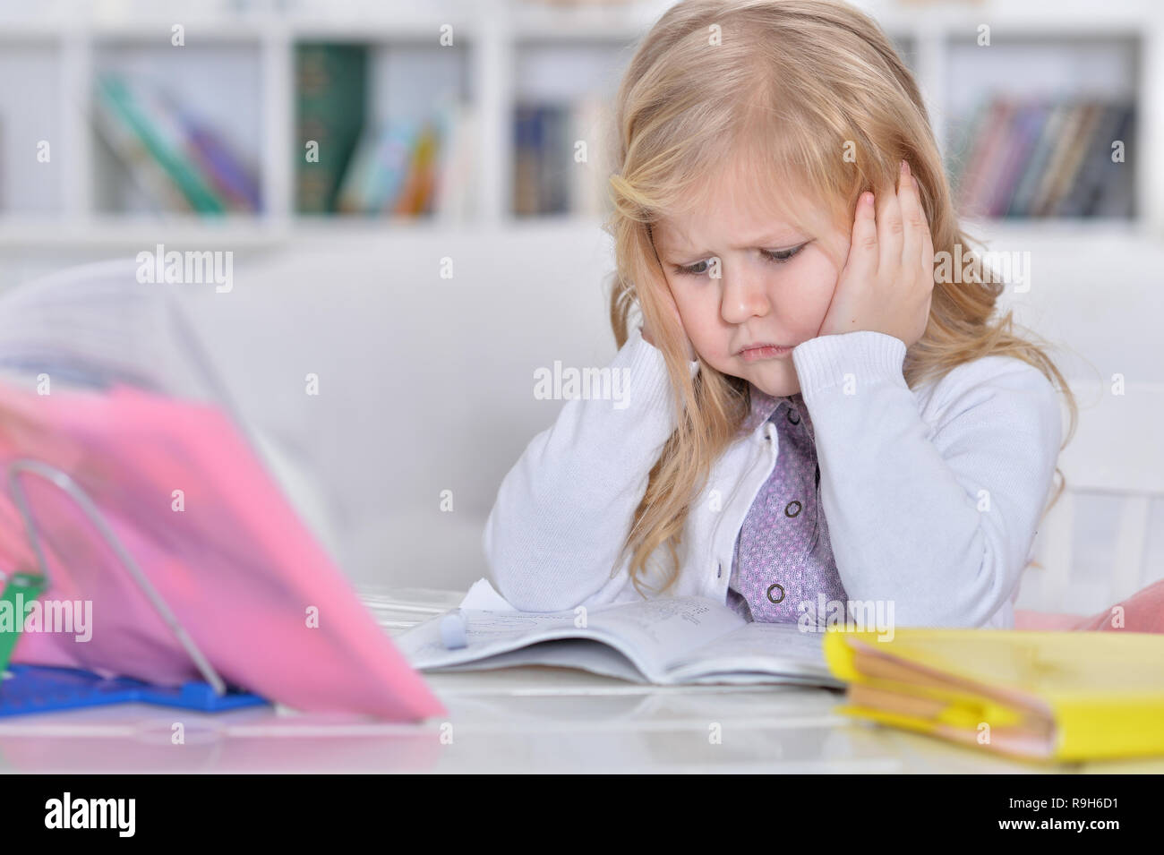 Cute tired schoolgirl sitting at table and doing homework at home Stock ...