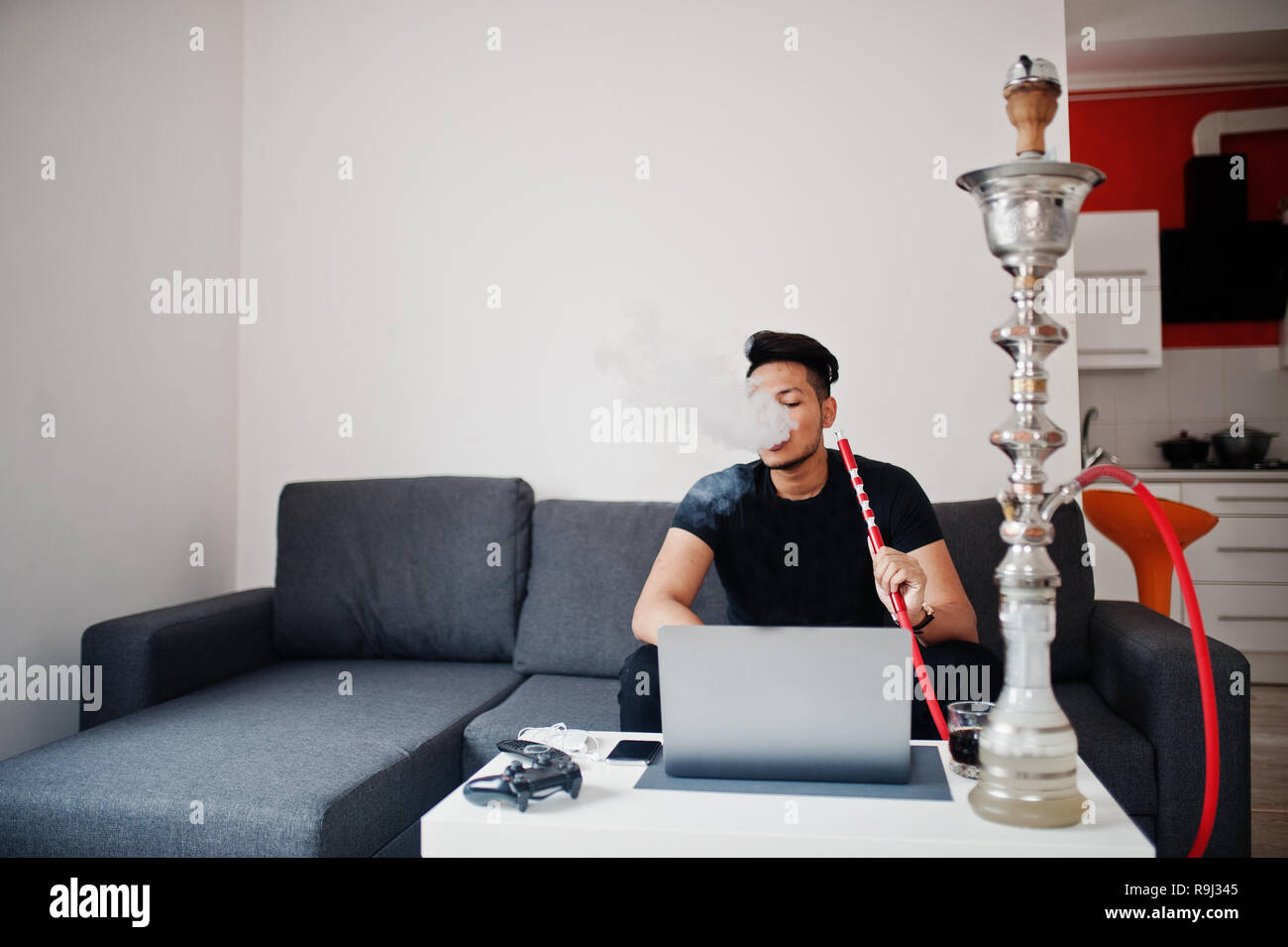 Handsome and fashionable indian man in black sitting at room, smoking ...