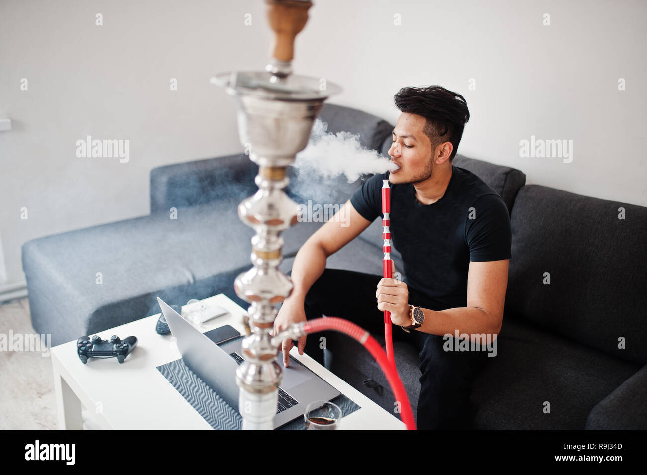Handsome and fashionable indian man in black sitting at room, smoking ...