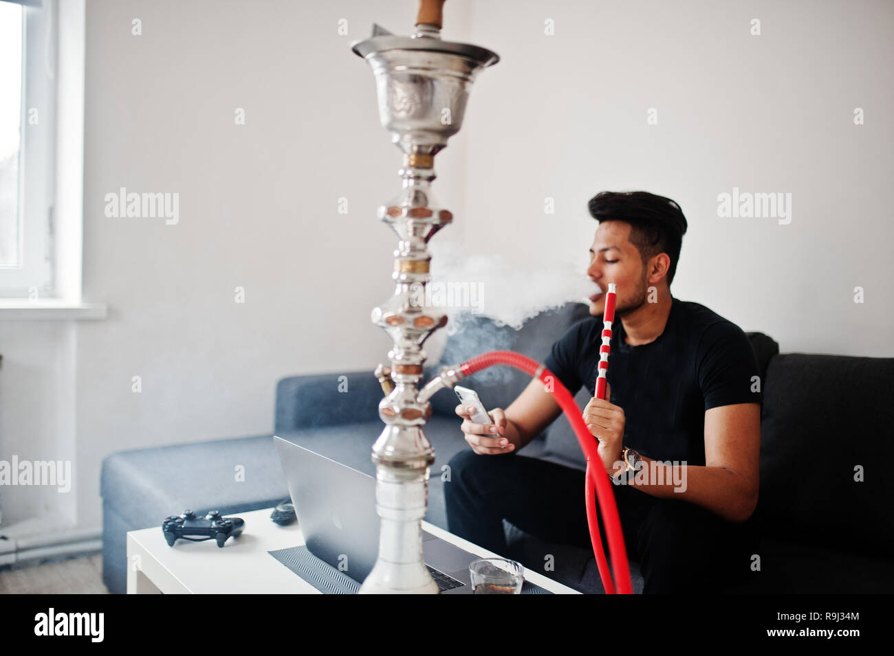 Handsome and fashionable indian man in black sitting at room, smoking ...