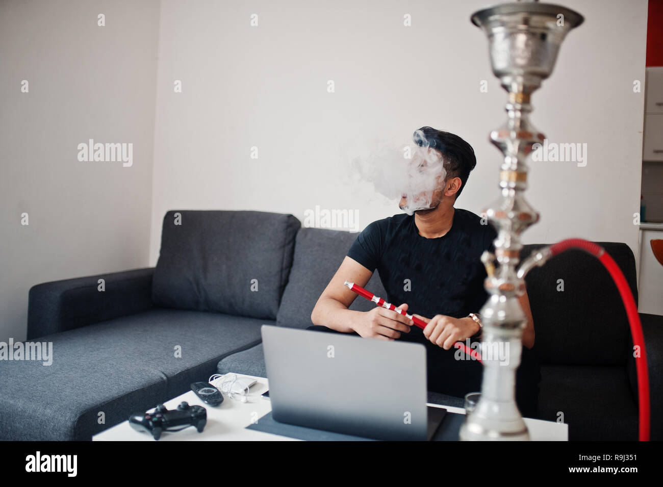 Handsome and fashionable indian man in black sitting at room, smoking ...