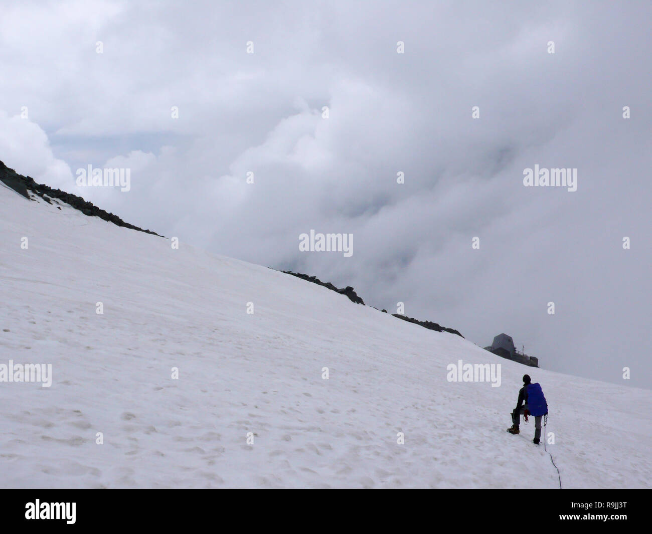 mountain climber hiking down over an alpine glacier with a mountain hut ...