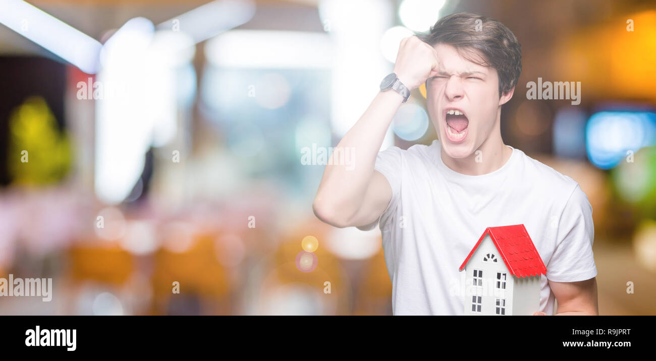 Young man holding house over isolated background annoyed and frustrated ...