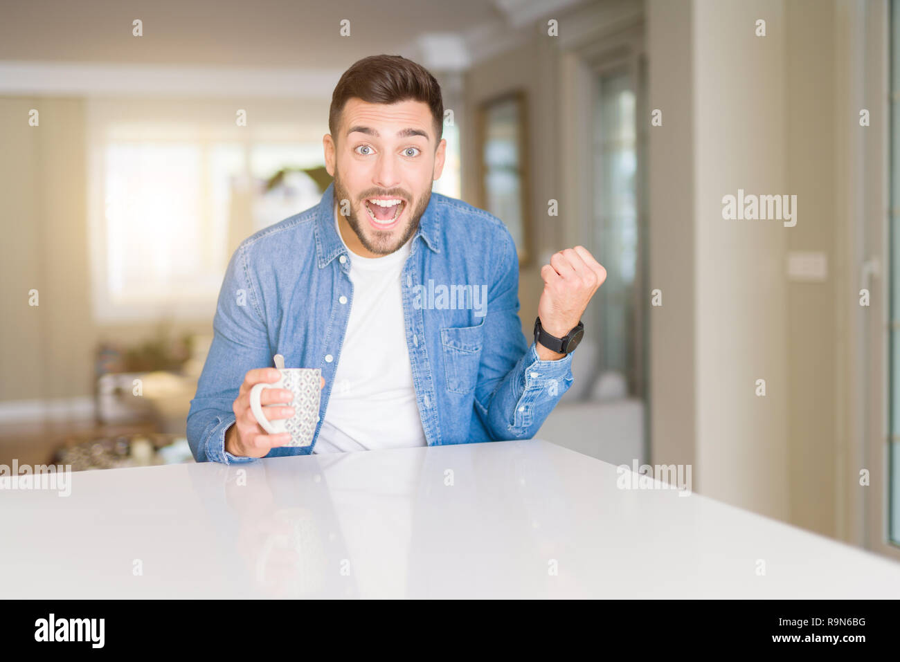Young handsome man drinking a cup of coffee at home screaming proud and ...