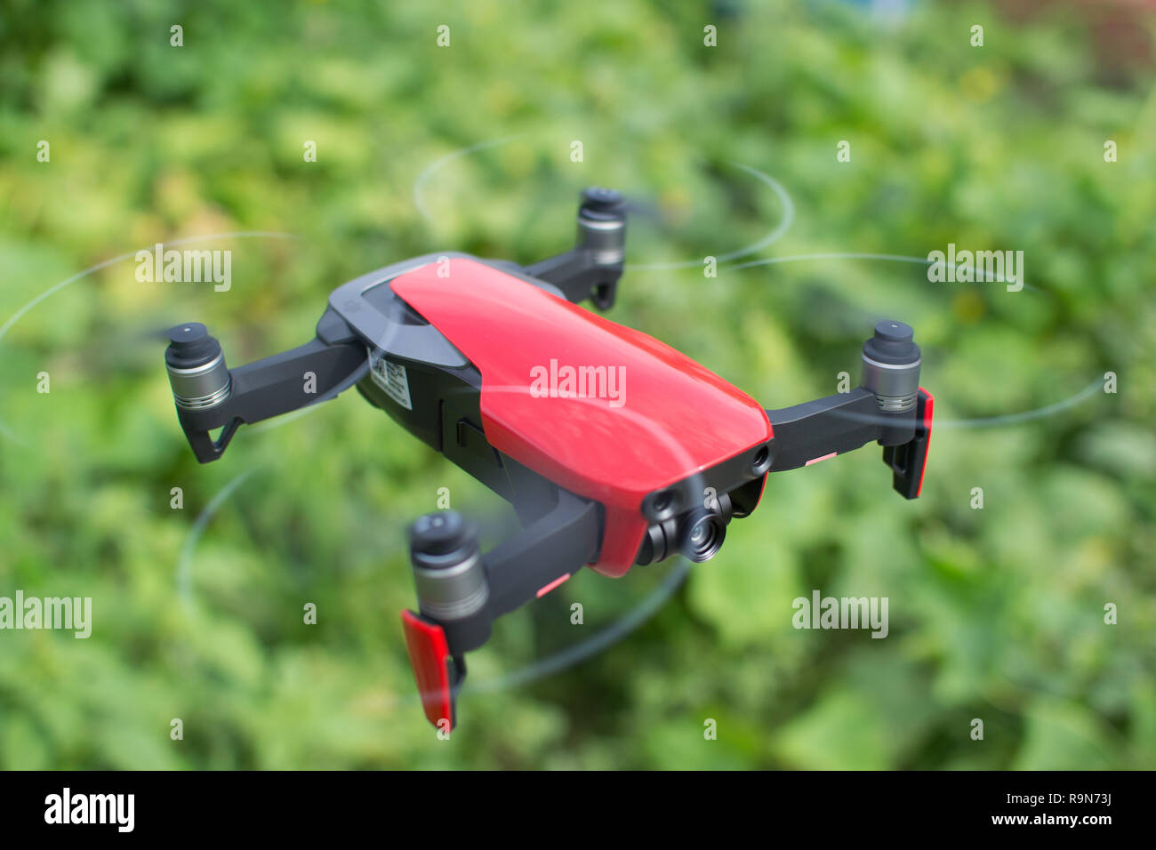 Small sized red drone is hovering in the air with green plants in the ...