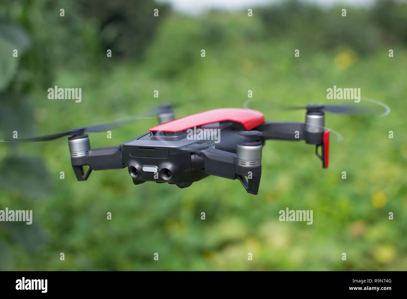 Small sized red drone is hovering in the air with green plants in the ...