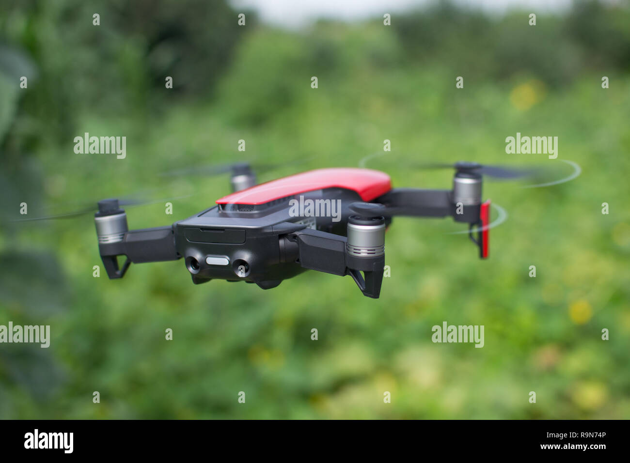 Small sized red drone is hovering in the air with green plants in the ...