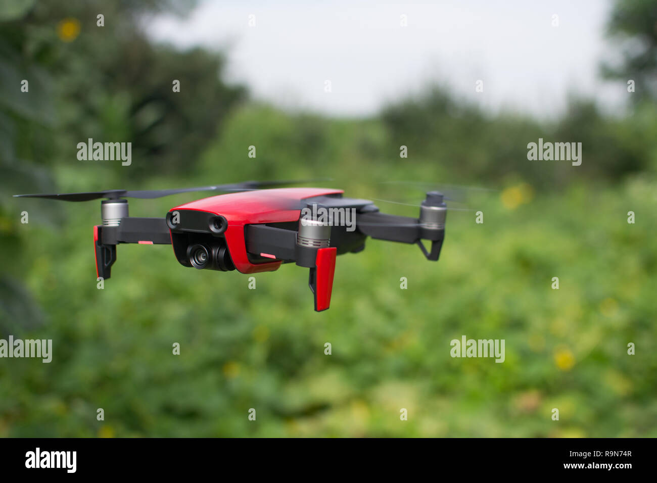 Small sized red drone is hovering in the air with green plants in the ...