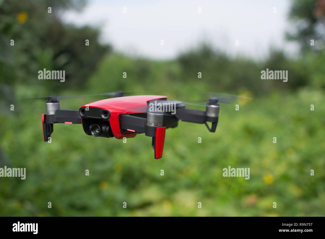 Small sized red drone is hovering in the air with green plants in the ...