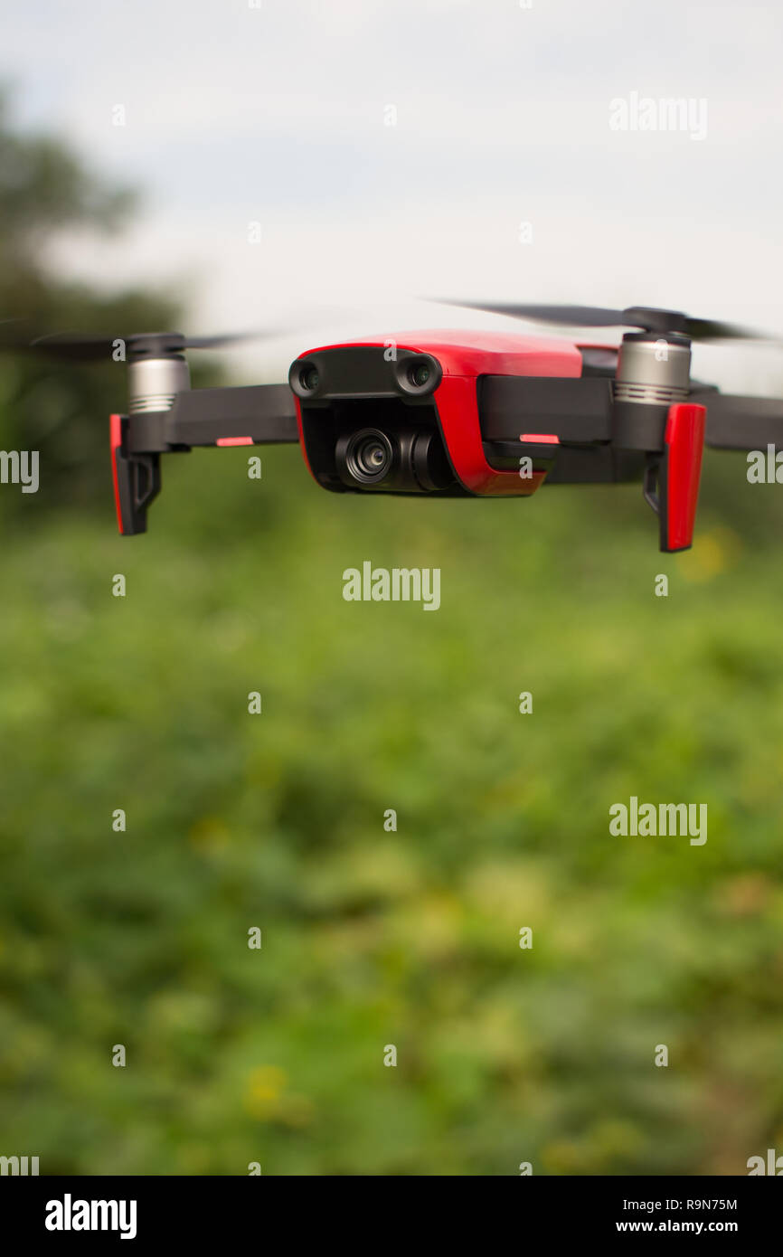 Small sized red drone is hovering in the air with green plants in the ...